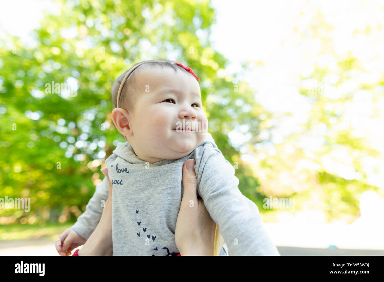 Baby girl being held up outdoors in front of tree looking to the right ...