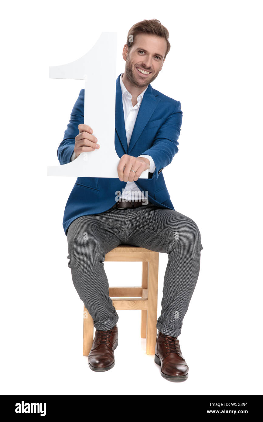 Casual man holding a number one with both hands while sitting and ...