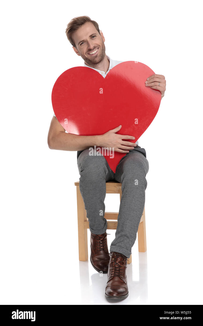 happy young casual man hugs a big red heart while sitting on chair on ...