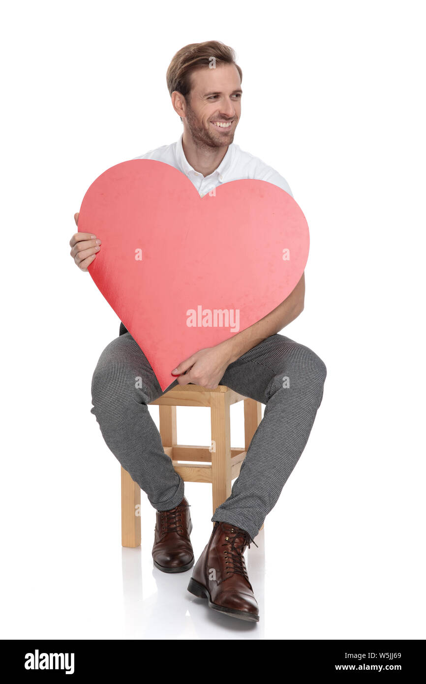 seated casual man holds big red heart and looks to side on white ...