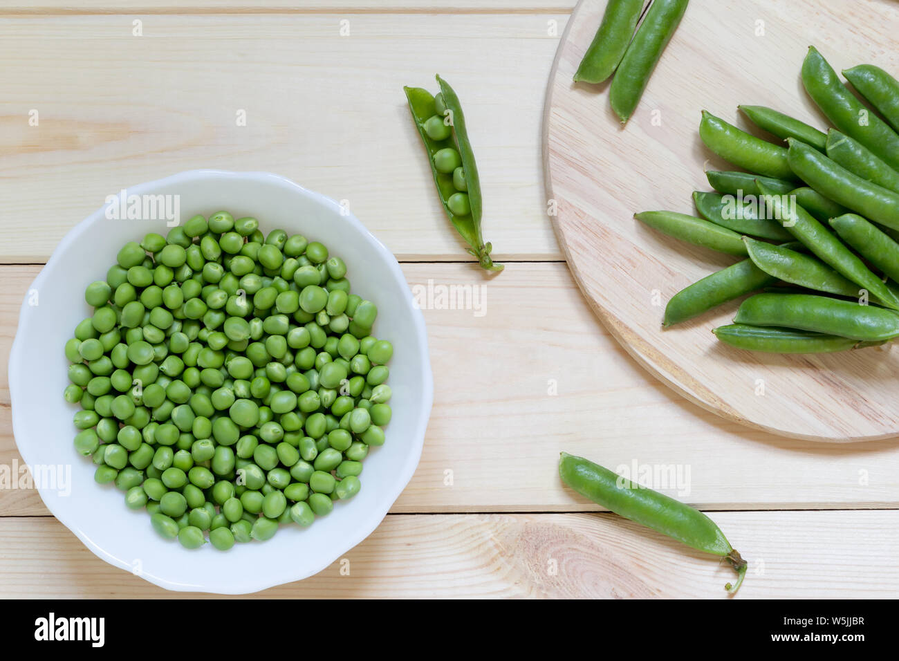Green peeled peas in white plate on light wooden table with whole pods ...