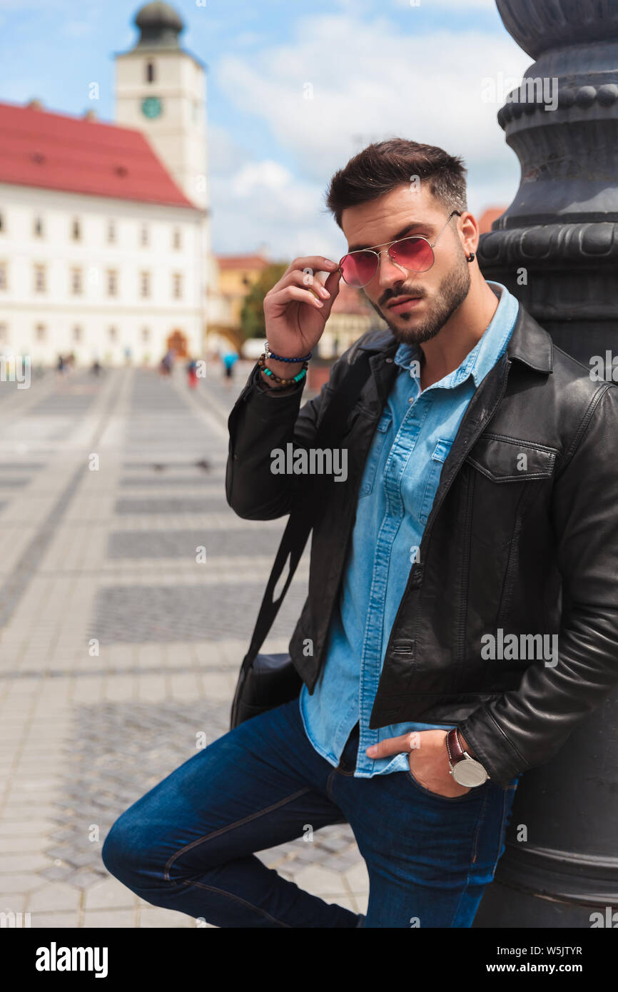 relaxed casual man leaning against lighting pole fixes sunglasses while ...