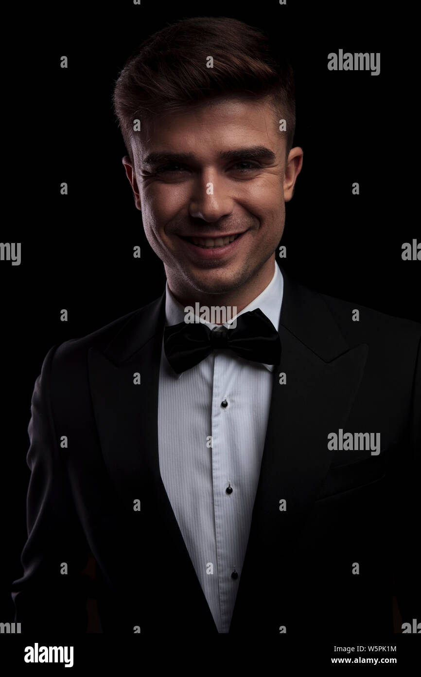 portrait of joyful young man in black tuxedo smiling while standing on ...