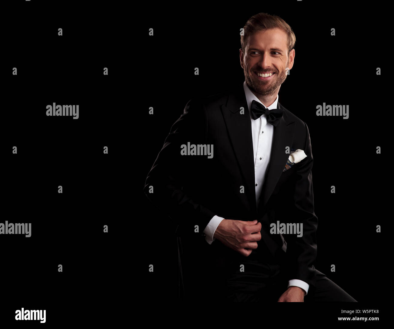 side view of a laughing elegant man in tuxedo holding button on black ...