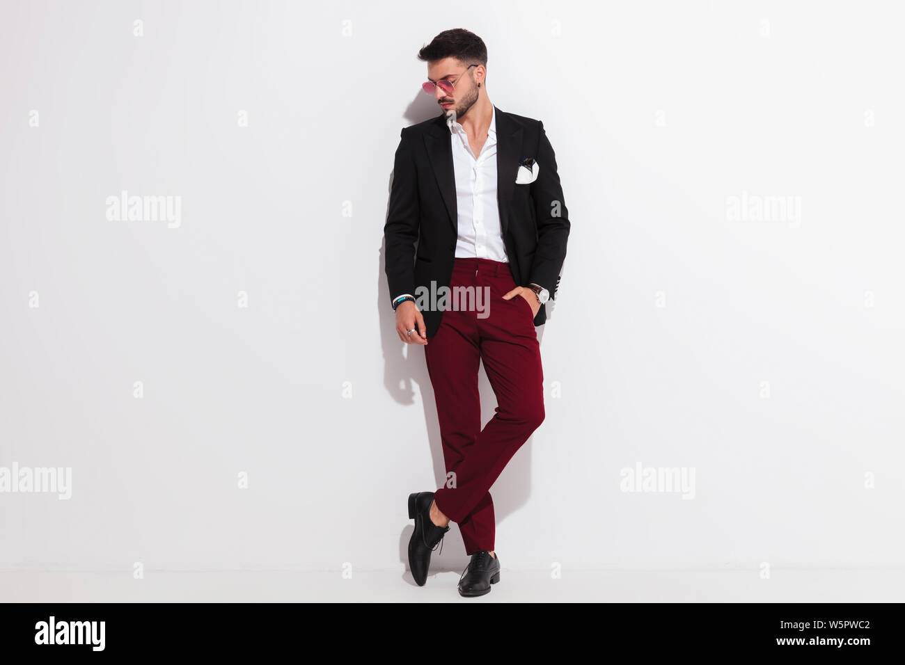 relaxed elegant man wearing a black suit is leaning against a white ...