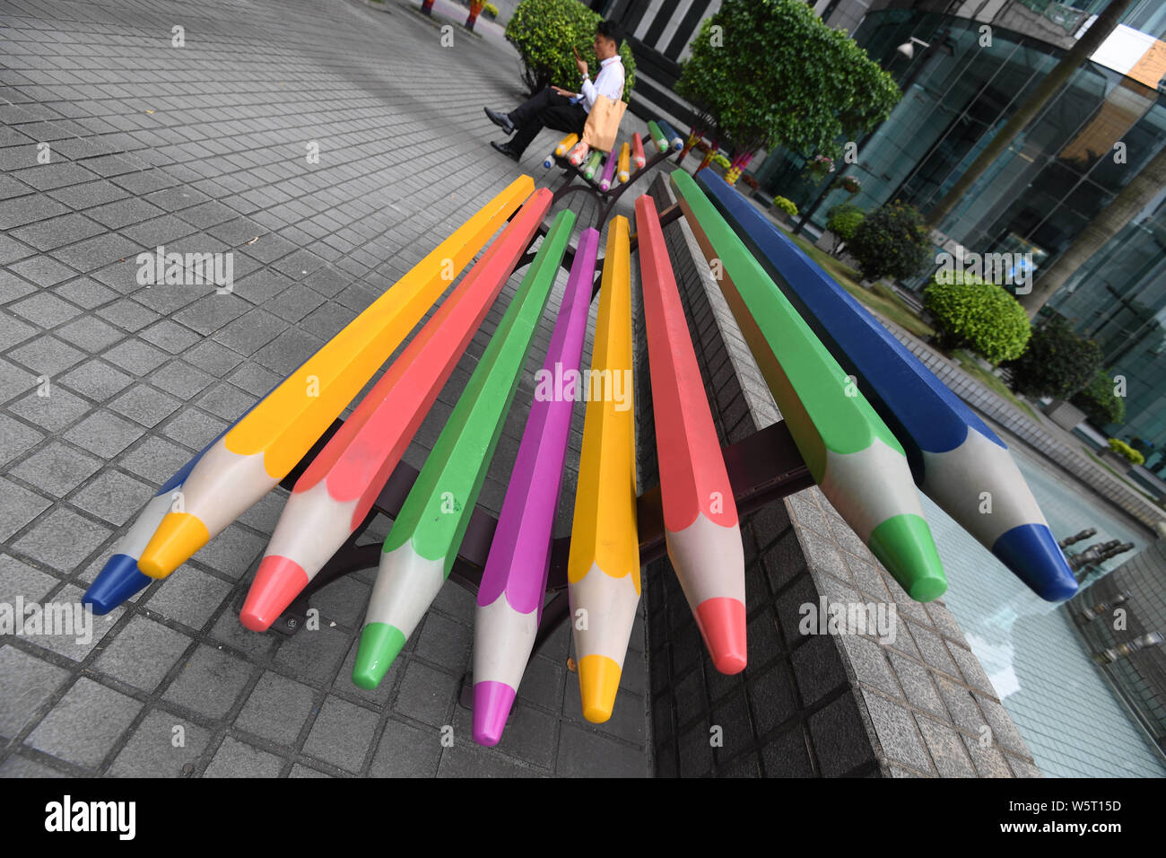 A Chinese man sits on a colored pencil bench next to others at a square ...