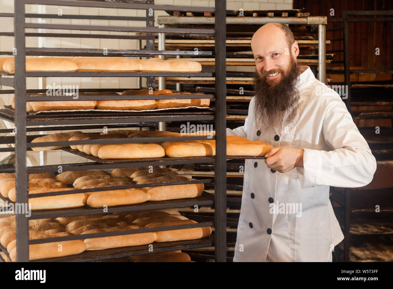 Portrait of pleasure young adult baker with long beard in white uniform ...