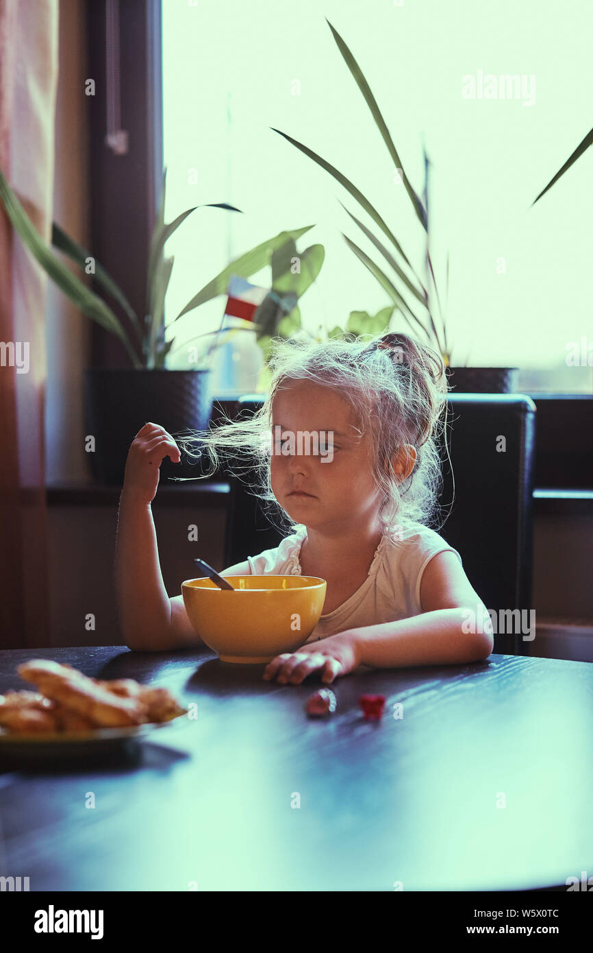 Little bored cute girl eating a breakfast, sitting by the table. Candid ...