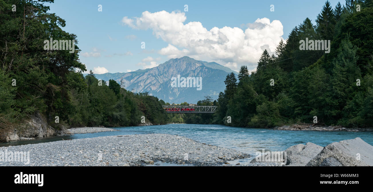 A red train crosses a bridge over the river Rhine in the Swiss Alps ...