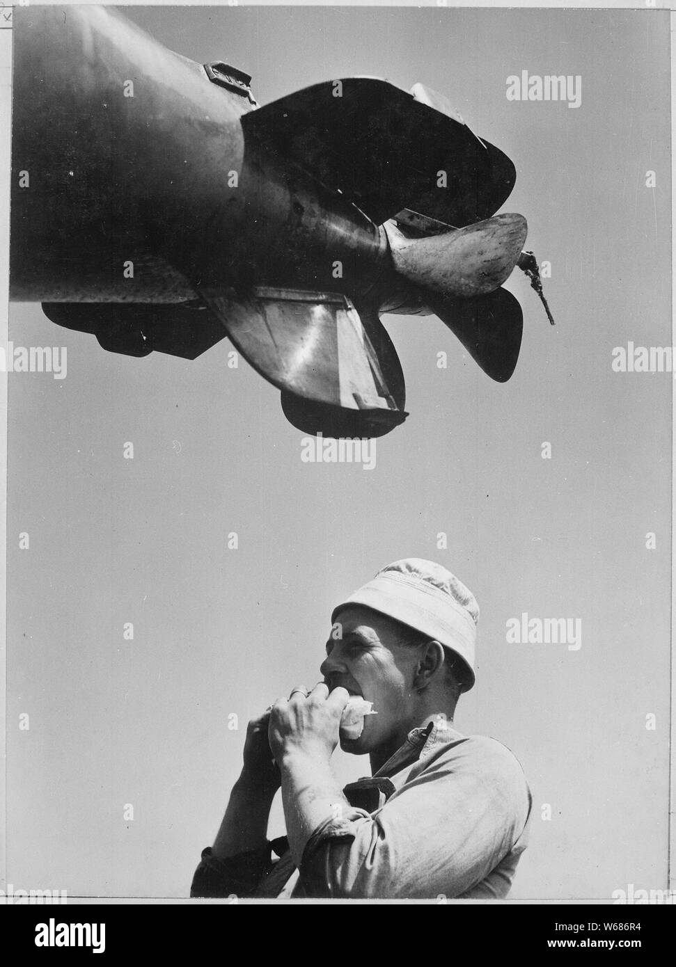 Sailor eating sandwich beneath propellers of torpedo being loaded ...
