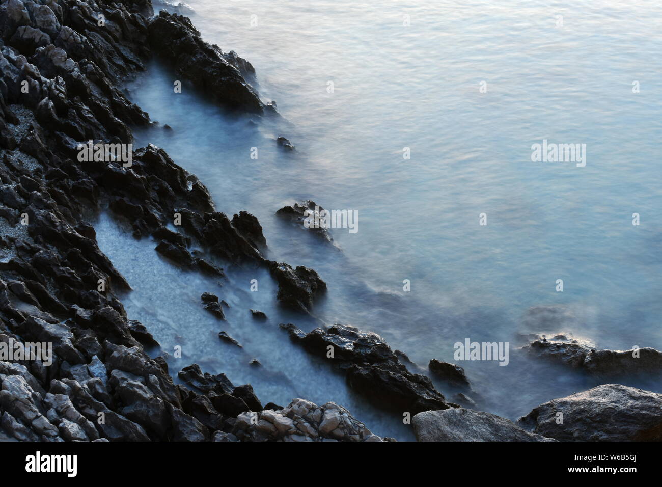 Rough dark rocky seashore with soft misty cloudy blue water. Visible ...