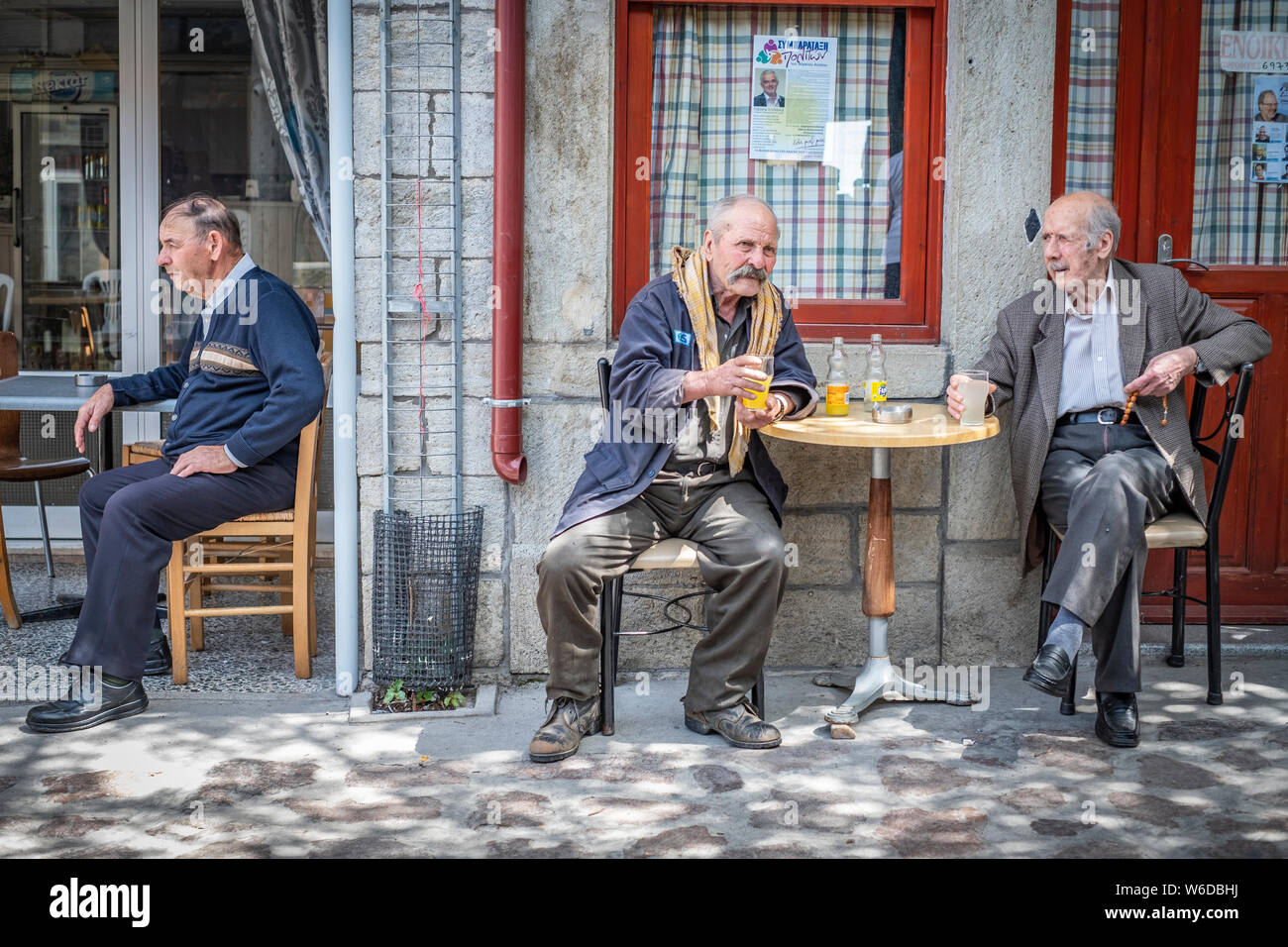 Three elderly Greek men sit in the shade of a village cafe observing ...