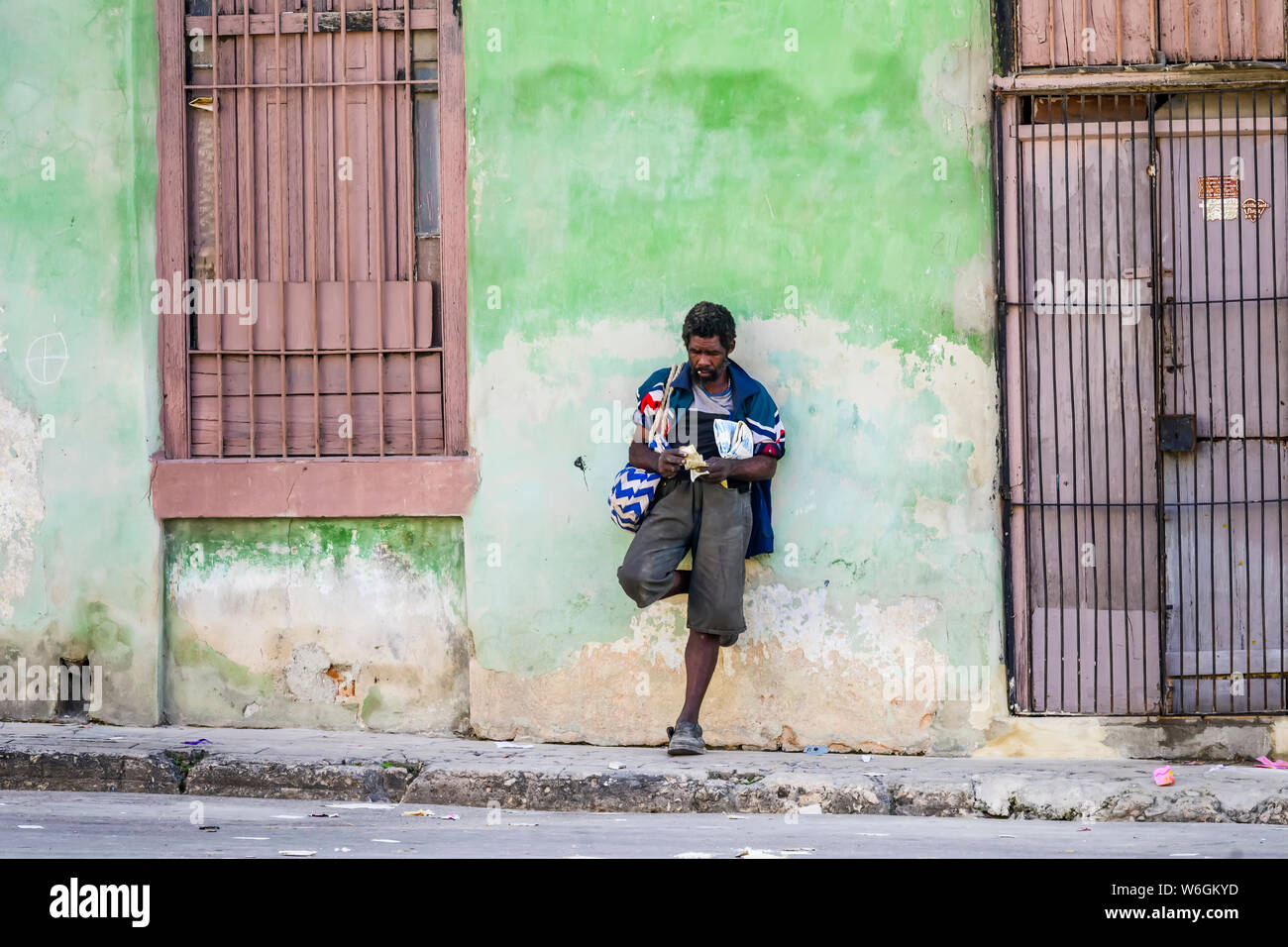 Cuban man stands leaning against the wall of a building along a street ...