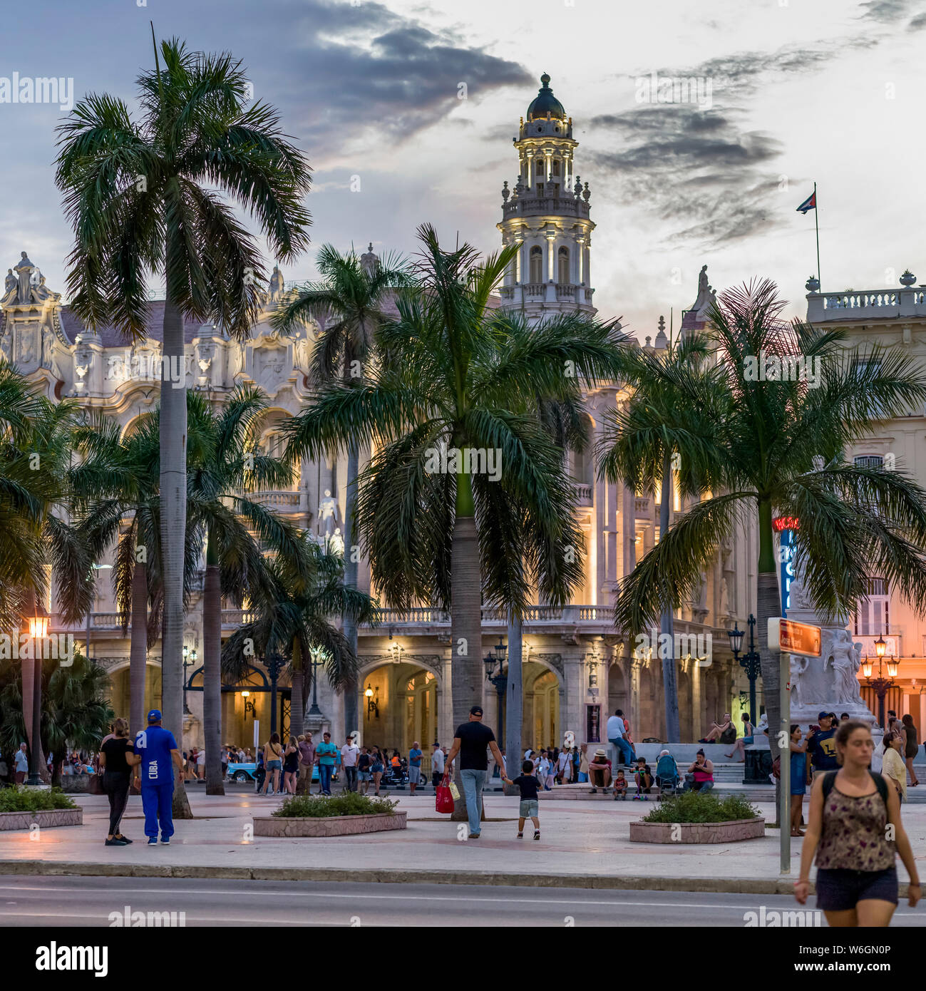 Pedestrians in a town square at dusk with palm trees and a tower in the ...