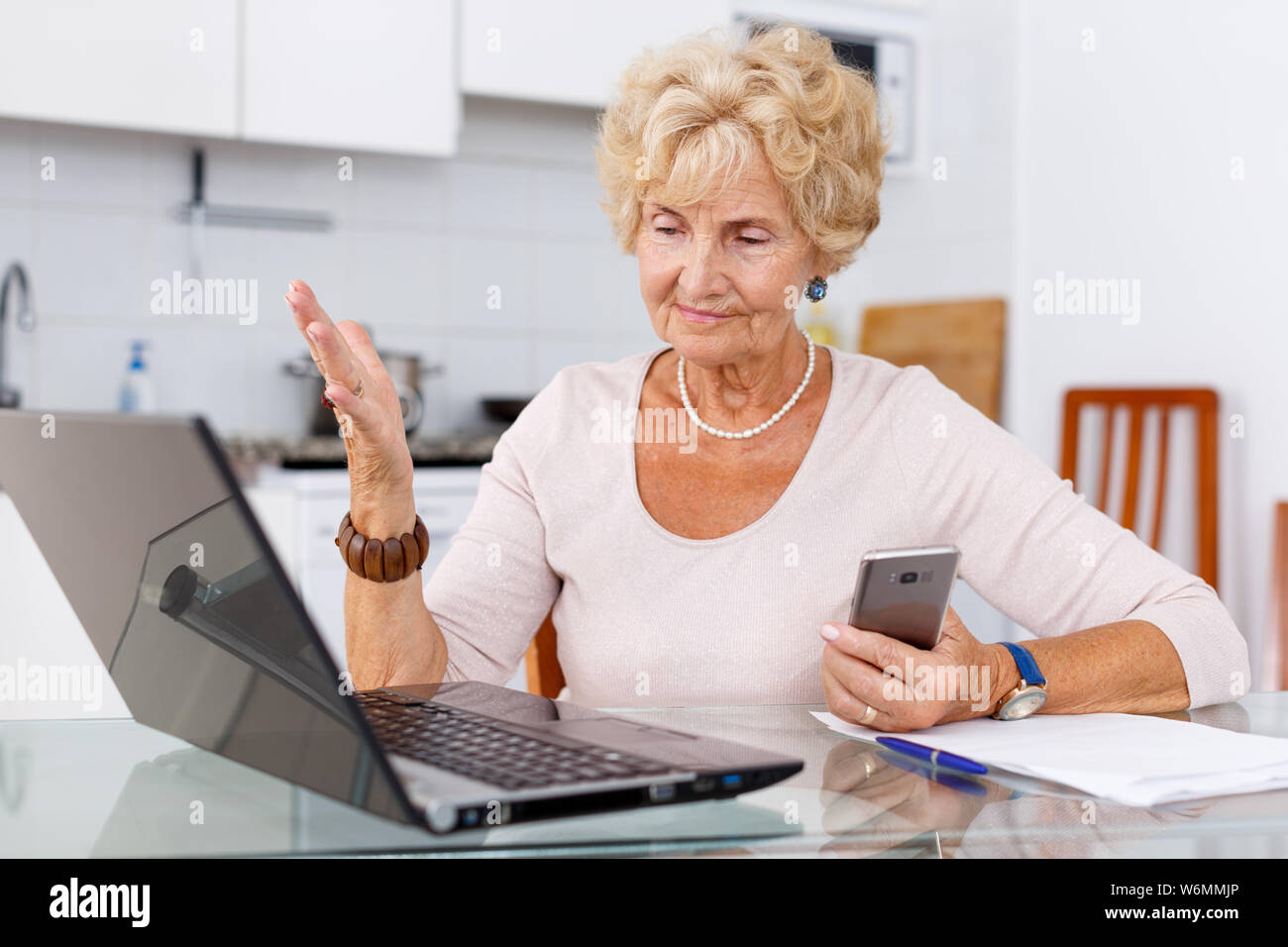Confused mature woman sitting at kitchen table and using laptop and ...