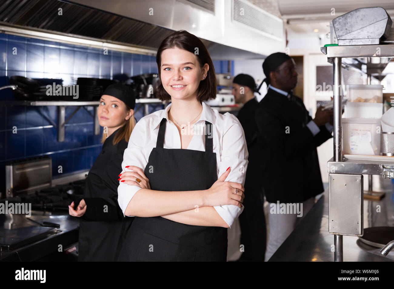 Confident young waitress standing with arms crossed in modern kitchen ...