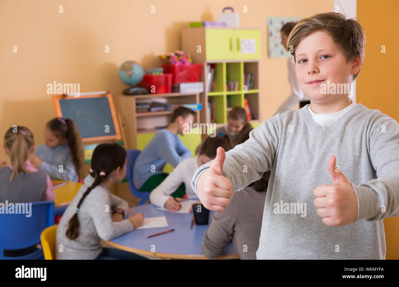 Smiling schoolboy hold thumbs up near children drawing at class Stock ...