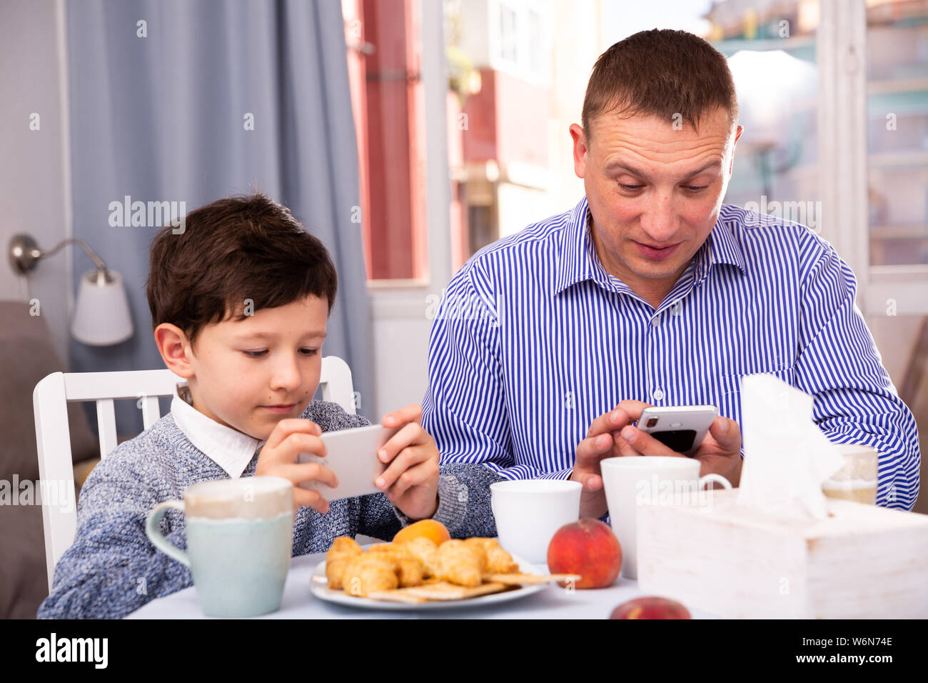 Cheerful man and his preteen son absorbedly looking at smartphones over ...