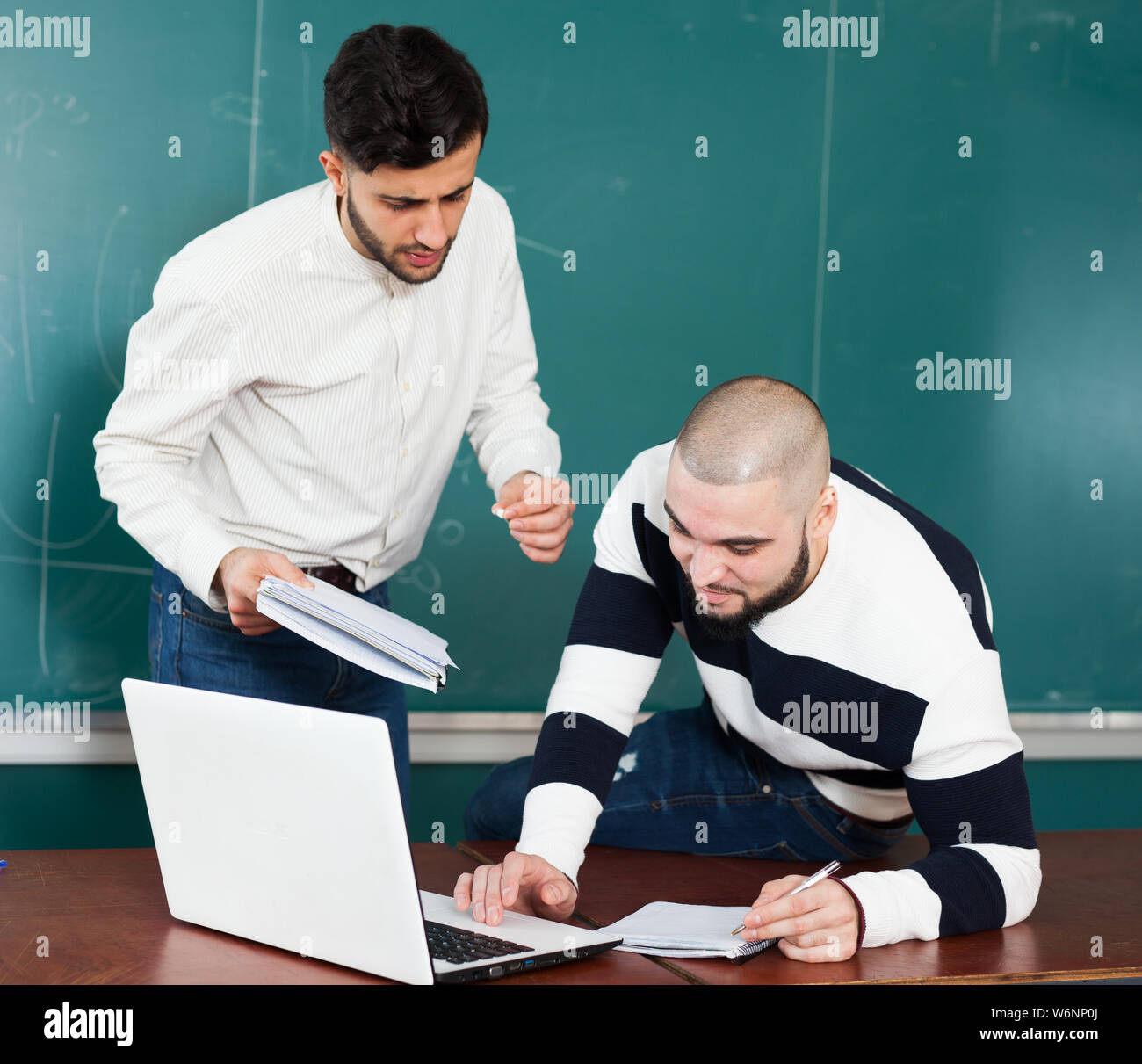 Two young handsome students working with laptop near blackboard at ...