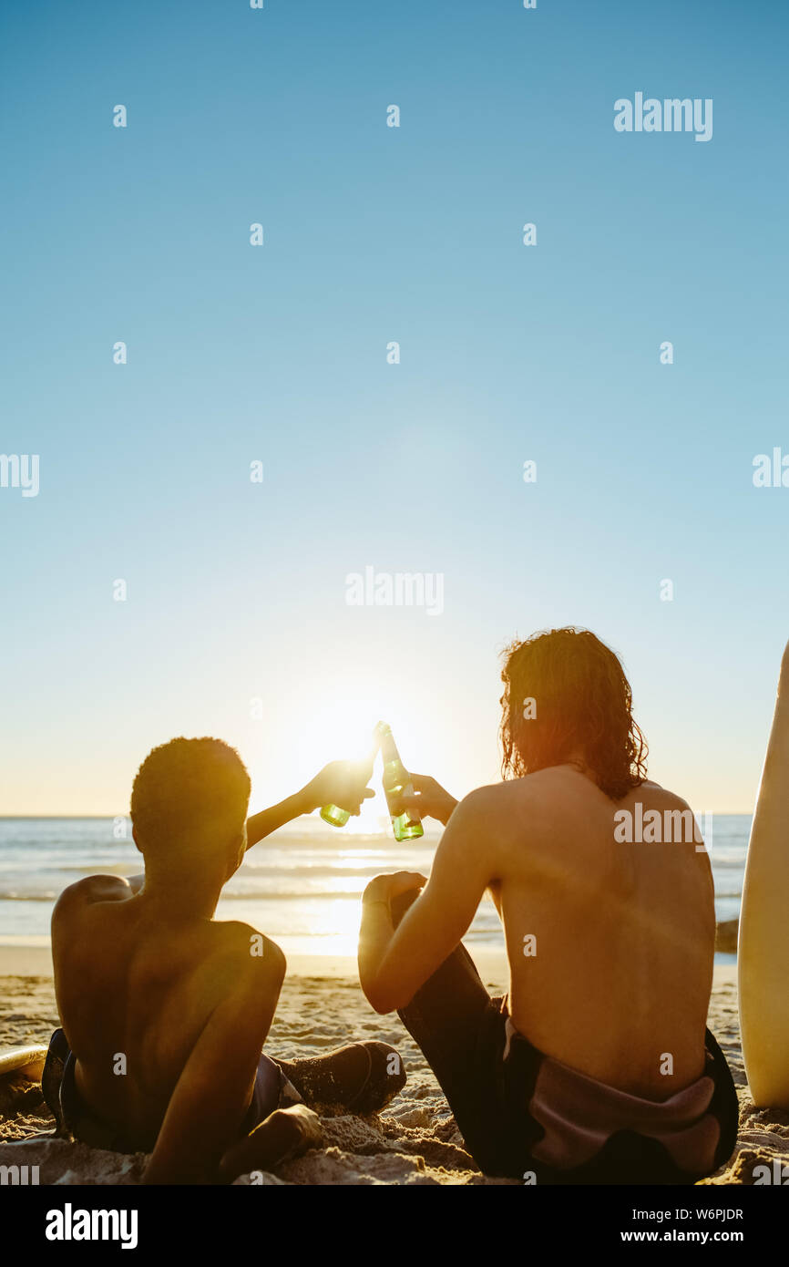 Rear view of two young men relaxing on the beach with beers. Young men ...