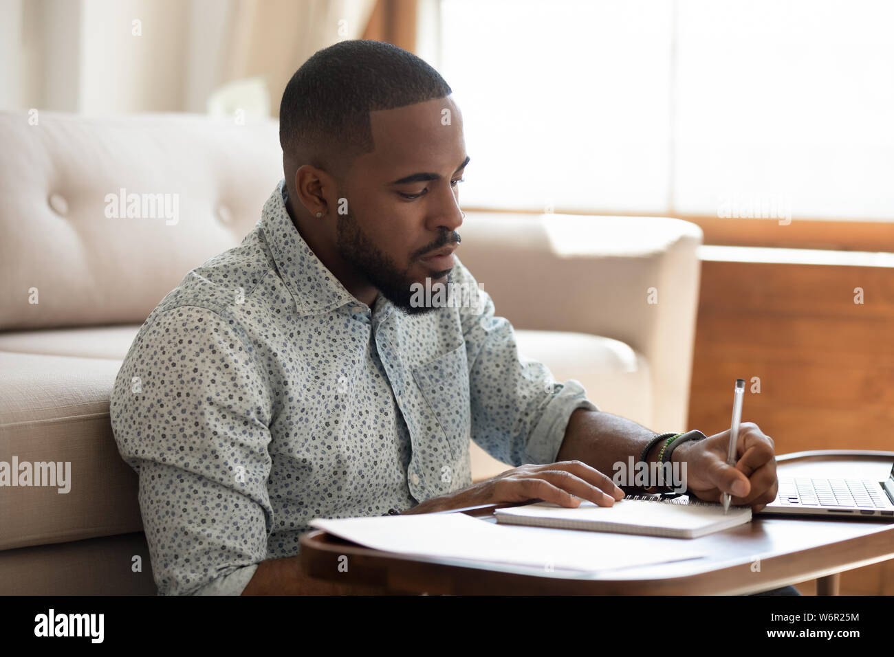Serious african student writing notes studying at home with laptop ...