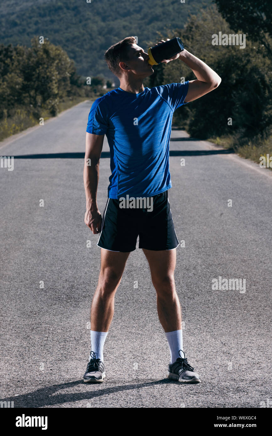 Fit male runner drinking water from a water bottle on an empty road in ...