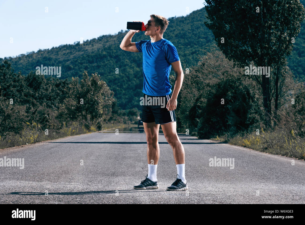 Fit male runner drinking water from a water bottle on an empty road in ...