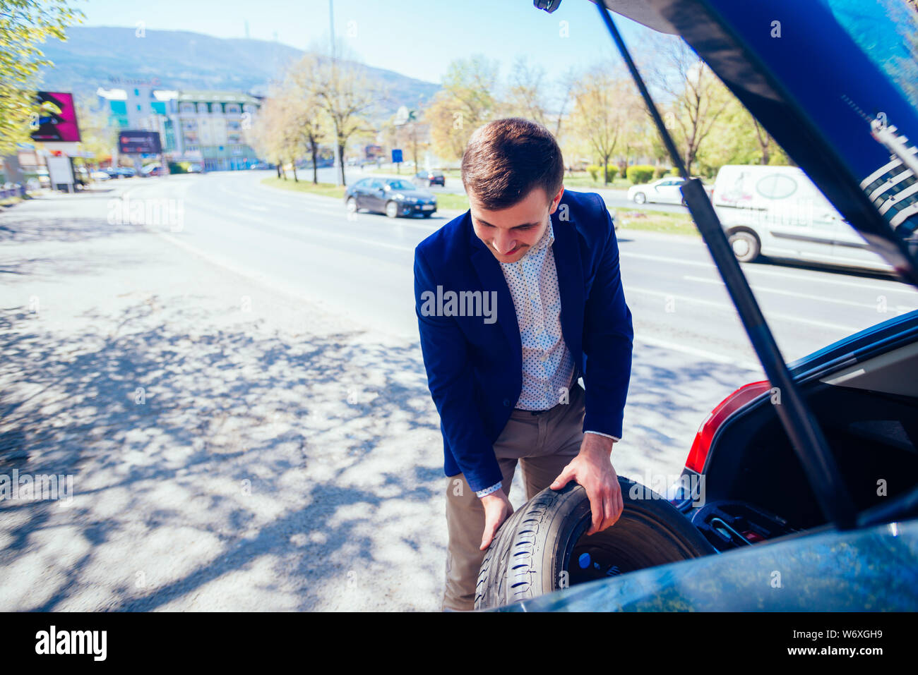 A tired businessman on a sunny day is taking a tire out of his car in ...