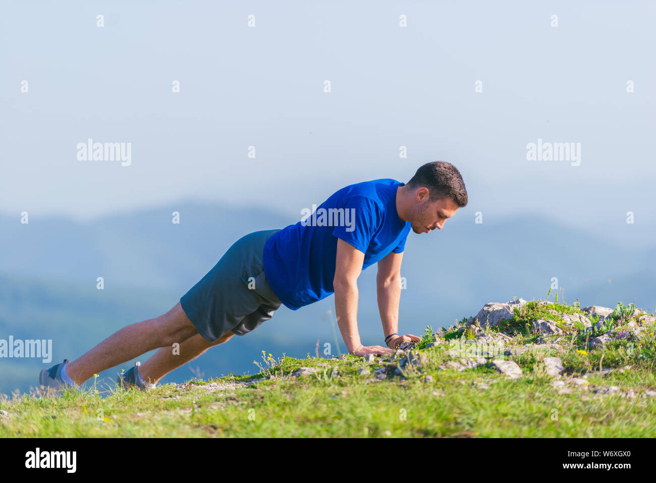 A young fit male athlete is doing push-ups outdoors on a cliff while ...