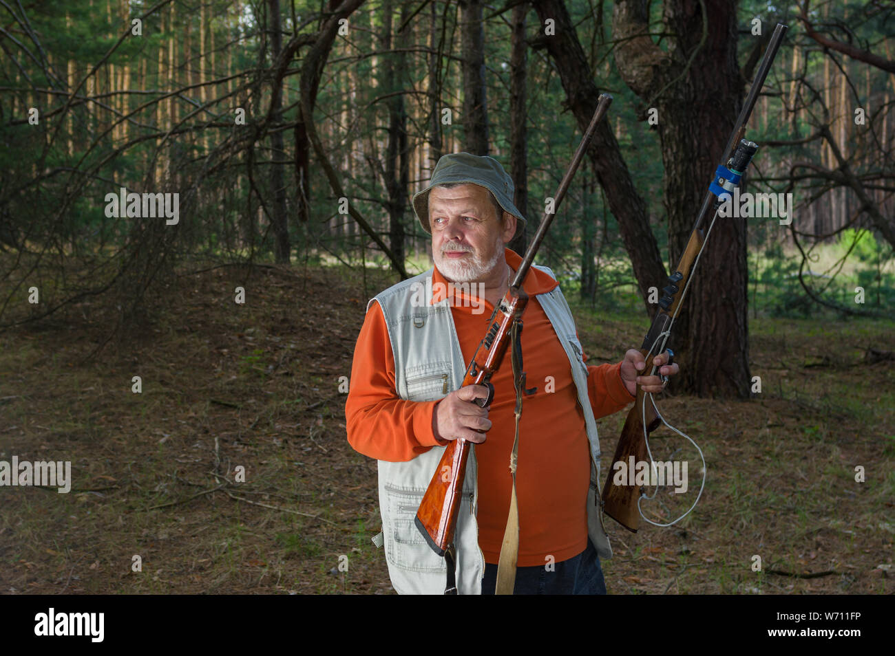 Portrait of senior ranger with two rifles standing in dark pine forest ...