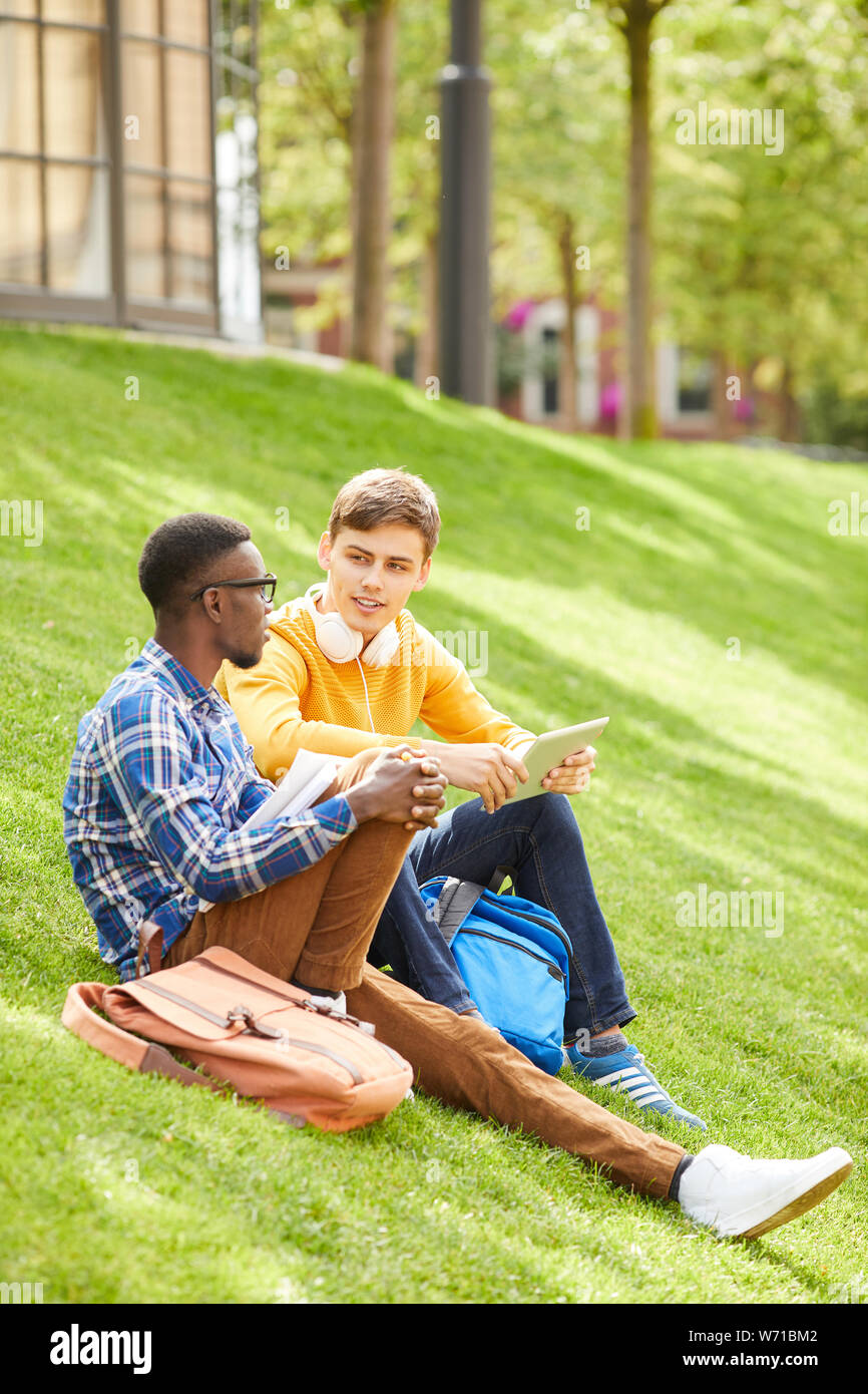 Side view portrait of two students sitting on green grass in campus and ...