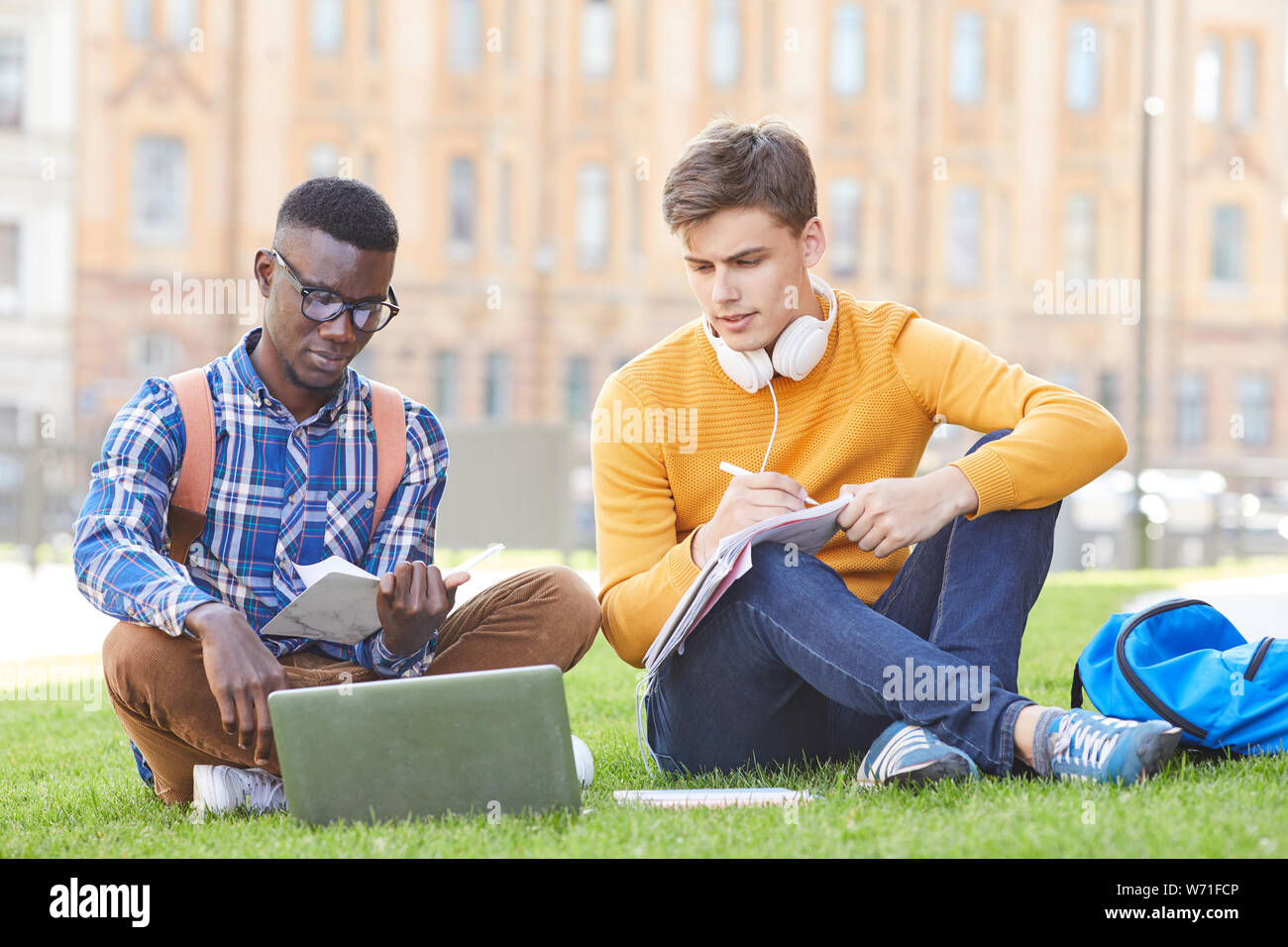Full length portrait of two college students studying outdoors sitting ...