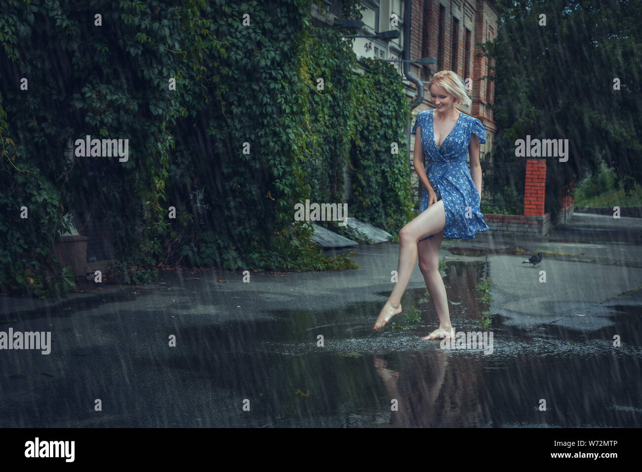 Young blonde woman in a dress frolics through puddles in the summer ...