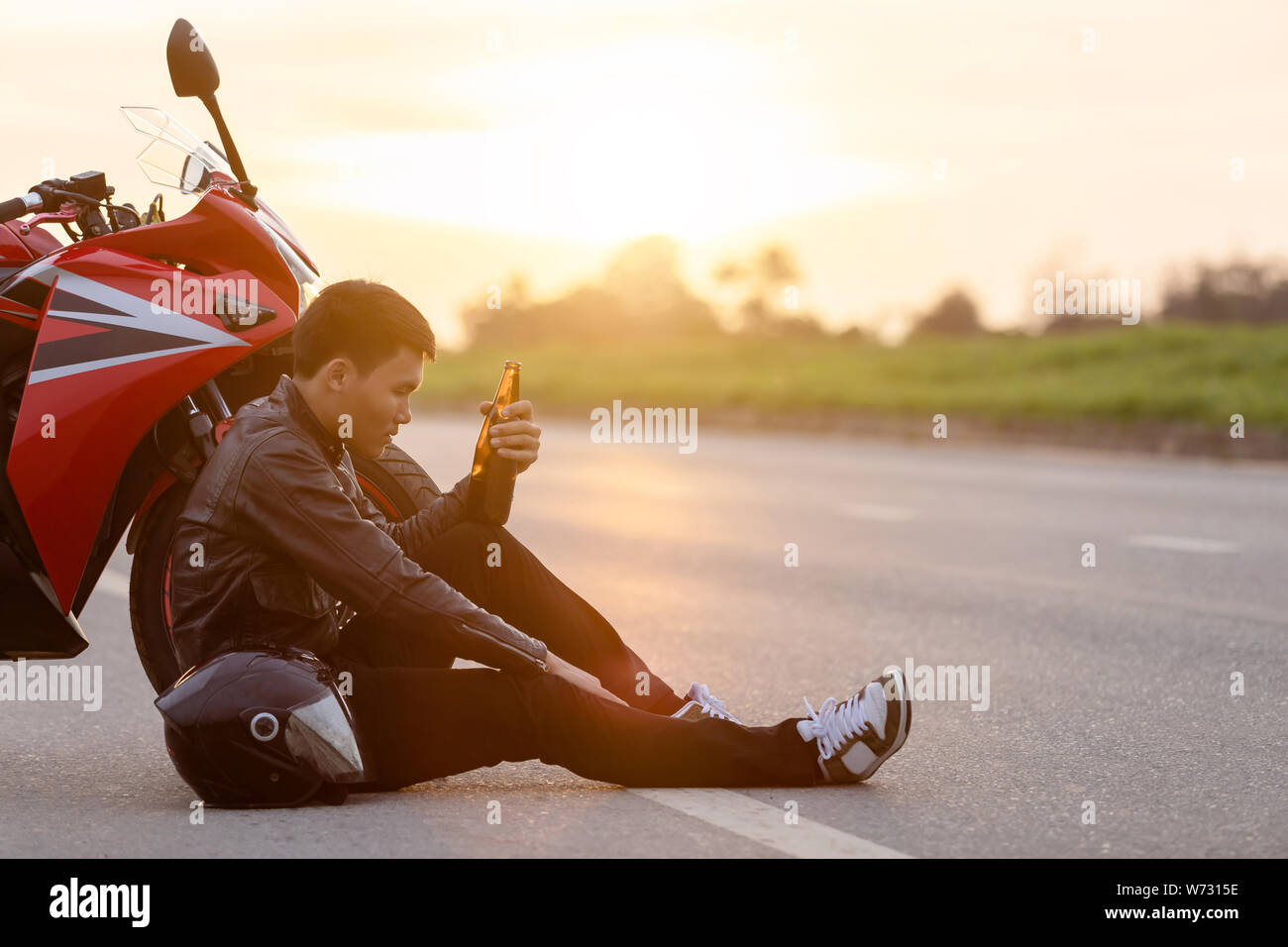 Motorcyclist sitting on the road beside his motorcycle and drinking an ...