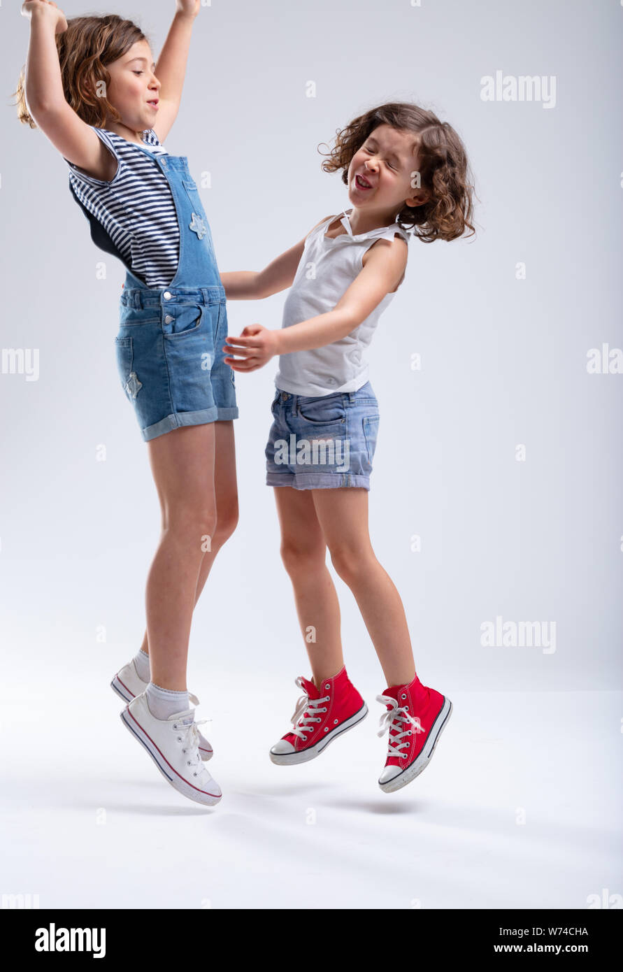 Two young sisters jumping and laughing together in blue denim shorts as ...