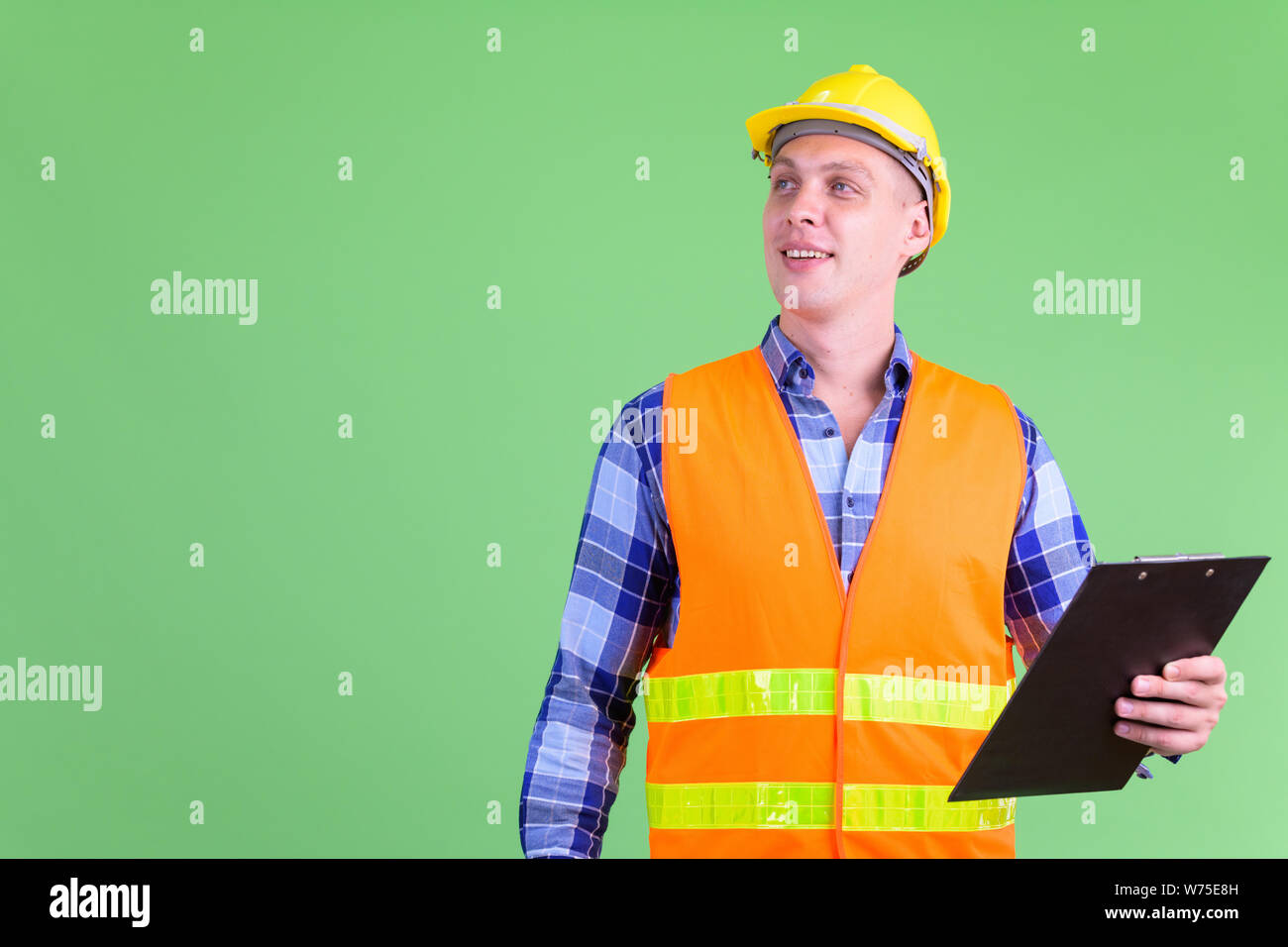 Happy young man construction worker thinking while holding clipboard ...