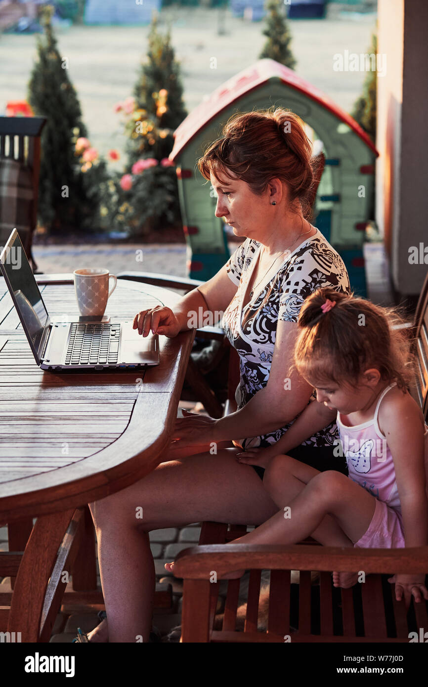 Mother working at home, using portable computer, sitting on patio with ...