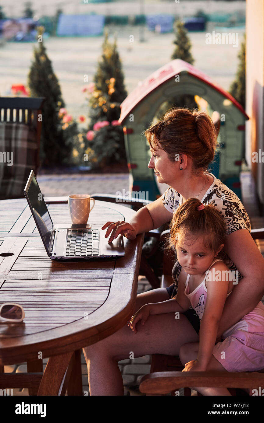 Mother working at home, using portable computer, sitting on patio with ...