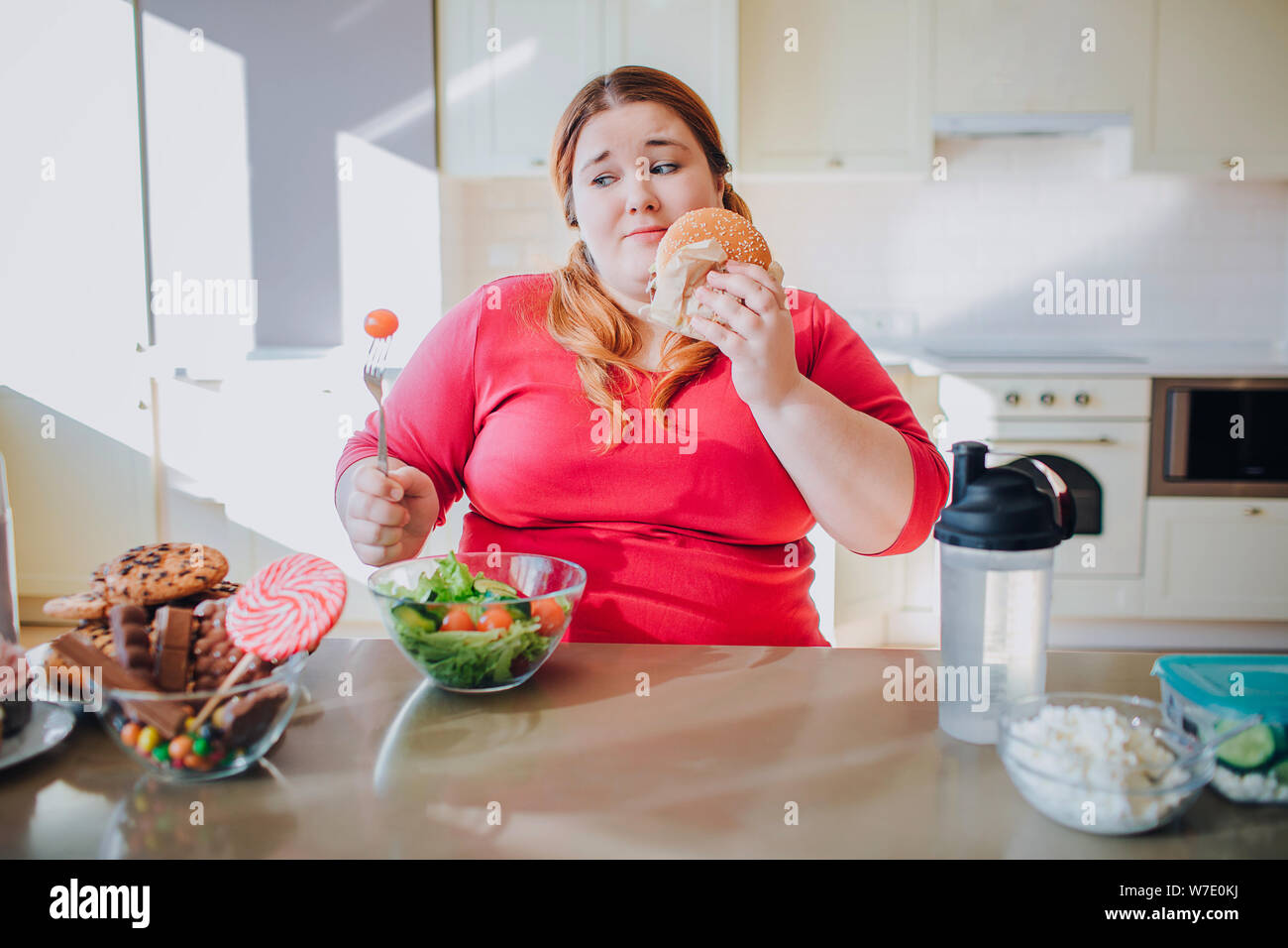 Fat young woman in kitchen sitting and eating junk food. Plus size ...