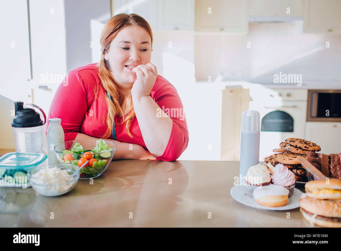 Fat young woman in kitchen sitting and eating food. Looking at sweet ...