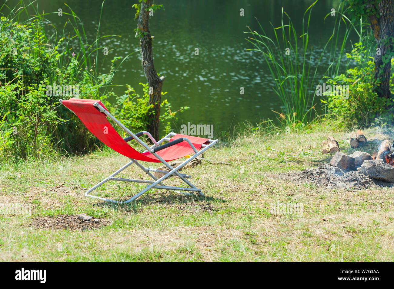 Empty red folding travel chair stands near bonfire on the lake coast at ...