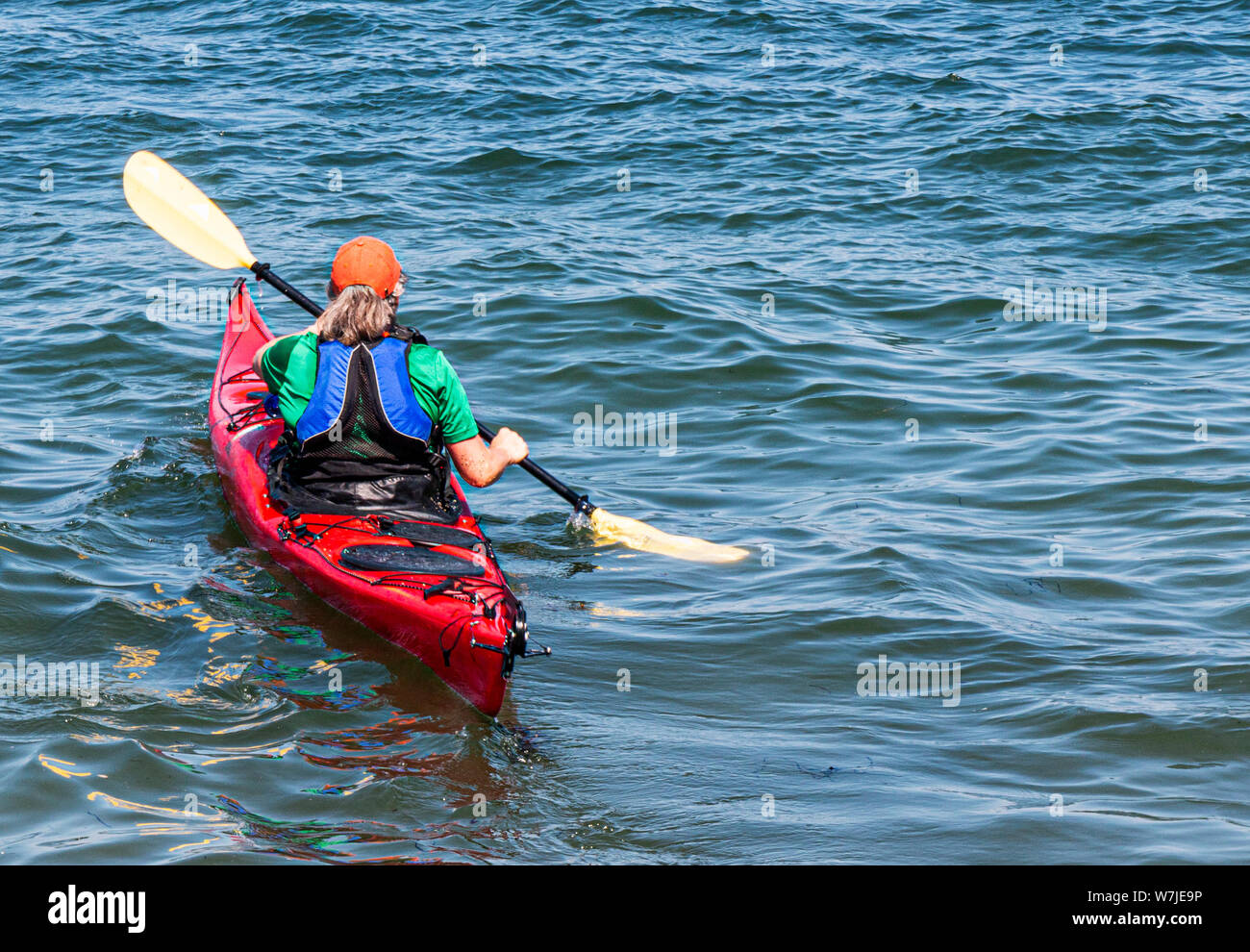 Rear view of a women in a red kayak paddling out into the bay wearing a ...