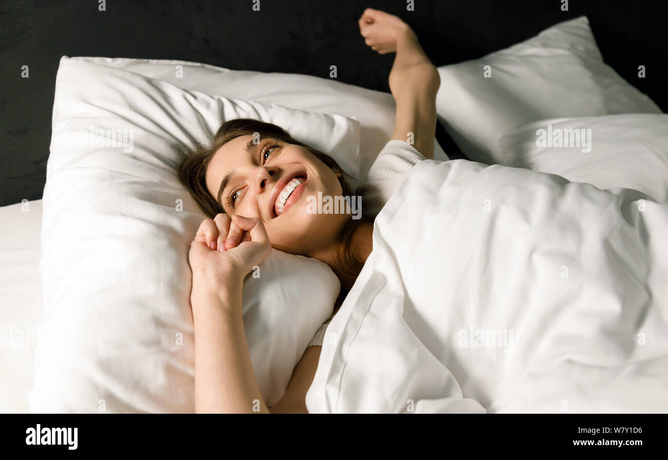 Beautiful brunette woman waking up in her white bed, she is smiling and ...
