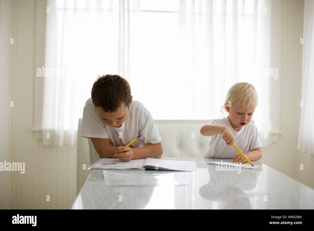 Young boy sitting at a dining table doing his homework with his younger ...