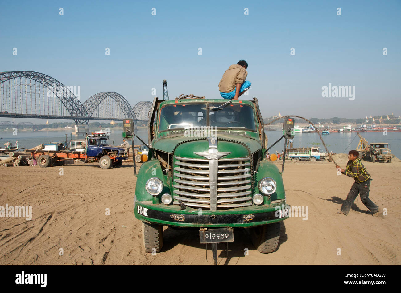 on the banks of the Irrawaddy river, trucks load tree trunks that will ...