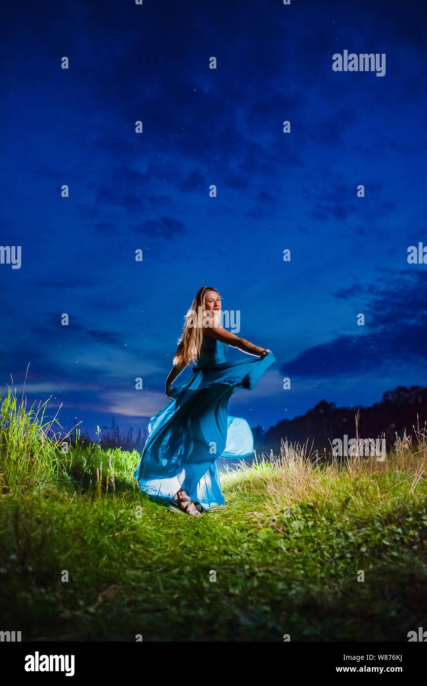 Beautiful blond model in long blue air dress posing in ray of light ...