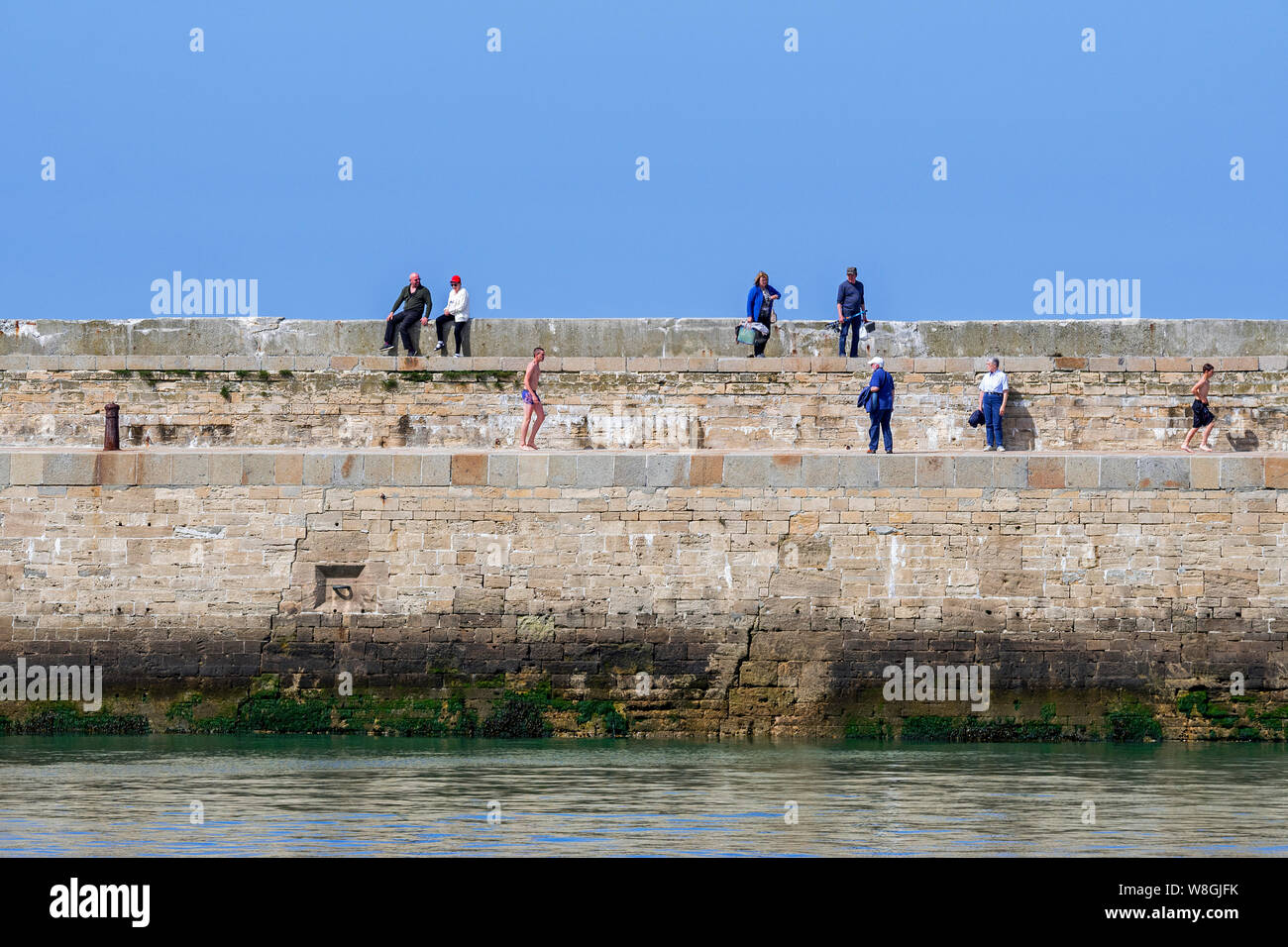 Tourists walking on old seawall / sea wall in the harbour at Port-en ...