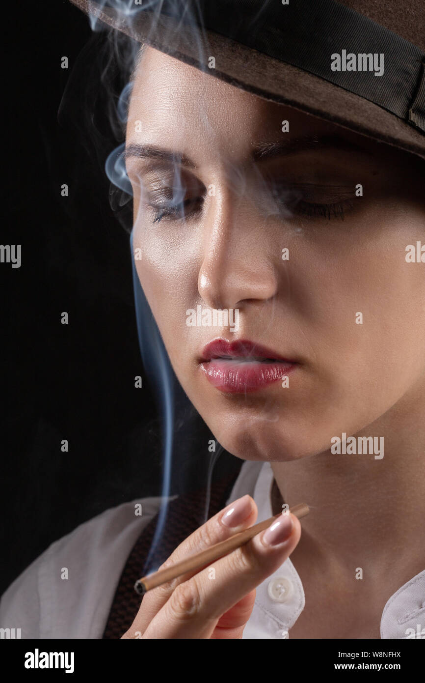 sad serious woman in hat smoking cigarette on black background, closeup ...
