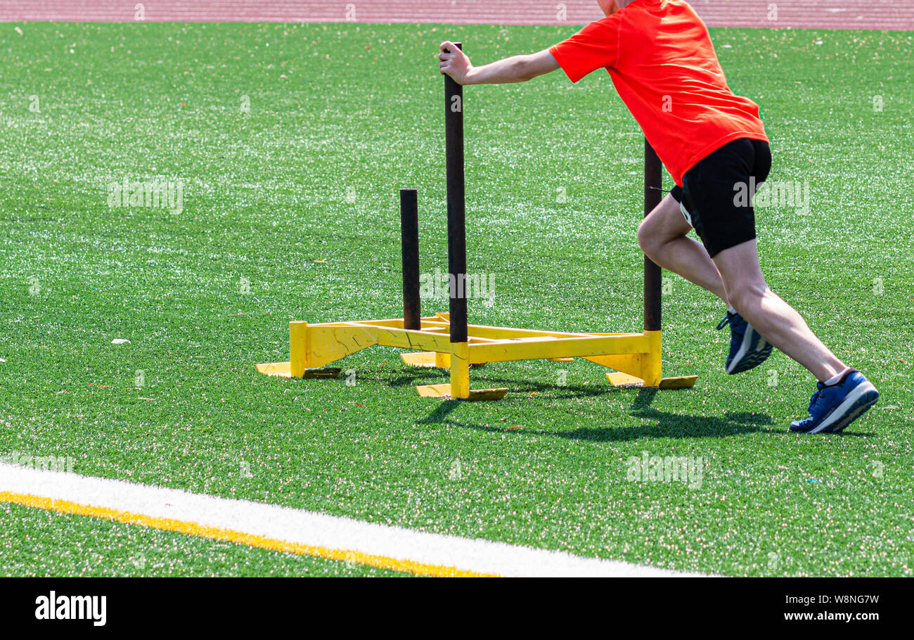 A young male runner is pushing a yellow sled on a green turf field at ...