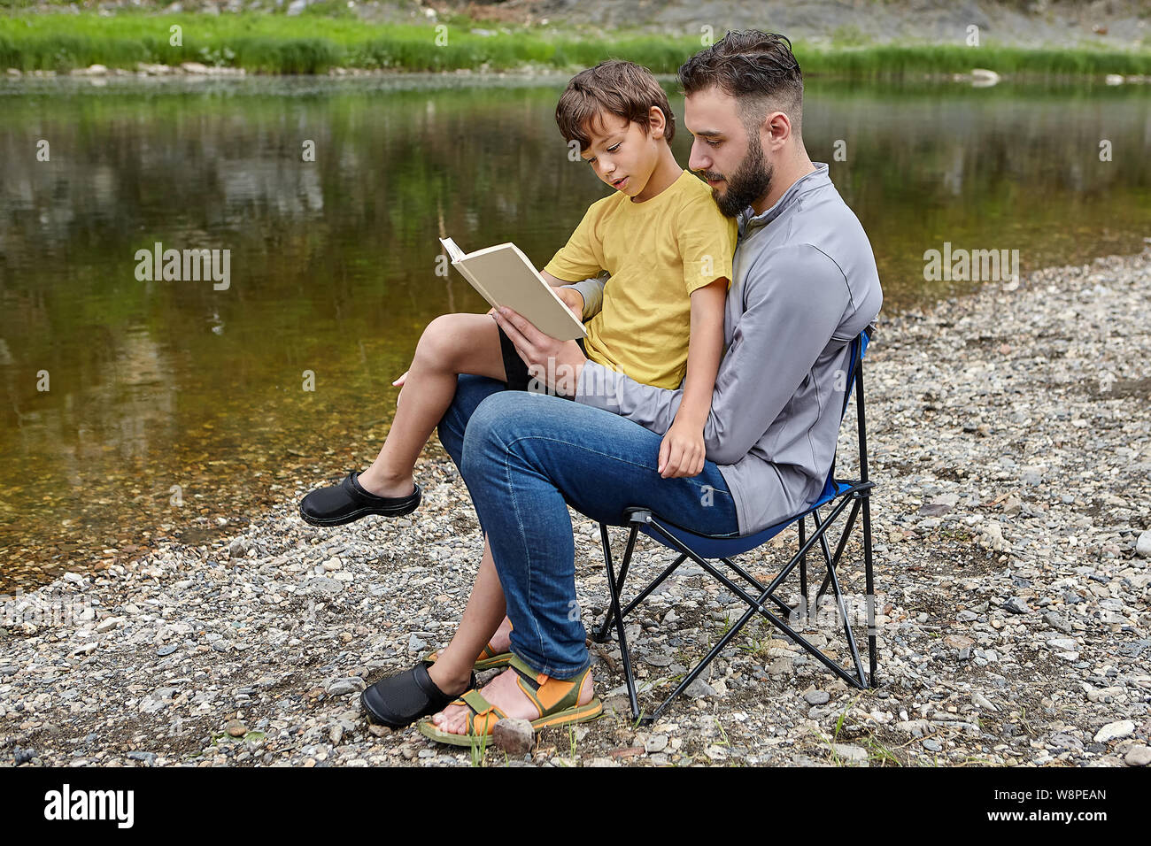 Father and son are sitting in a folding chair near the river, parent ...